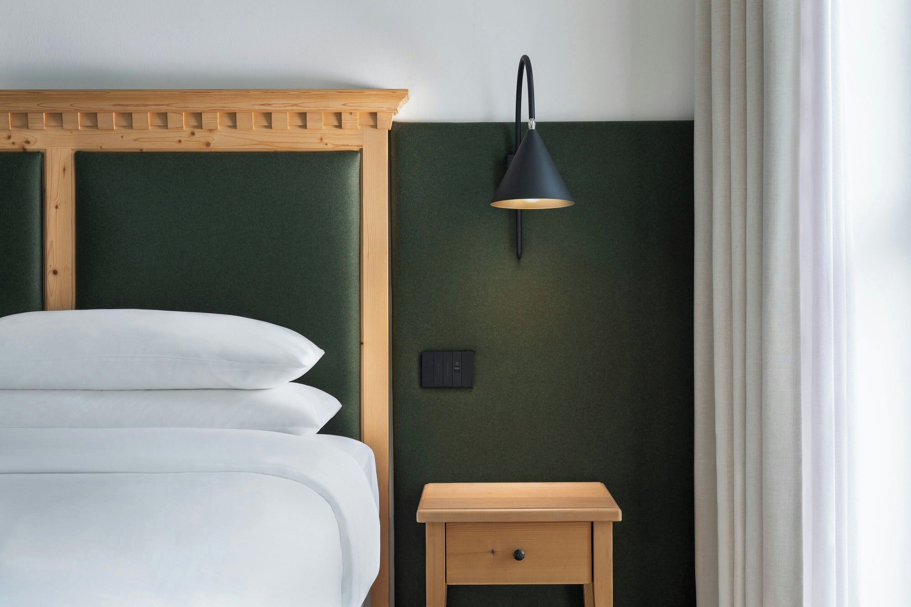 Uma cama com roupa de cama branca, cabeceira de madeira, mesa de cabeceira com gaveta e luminária fixada na parede.