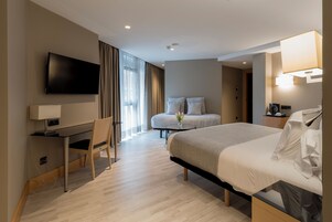 Quarto de hotel com cama, escrivaninha, cadeira, televisão e janela com cortinas.