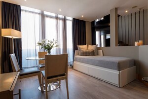 Quarto de hotel moderno com cama-sofá, mesa de jantar com cadeiras, um vaso com flores e uma janela grande com cortinas.