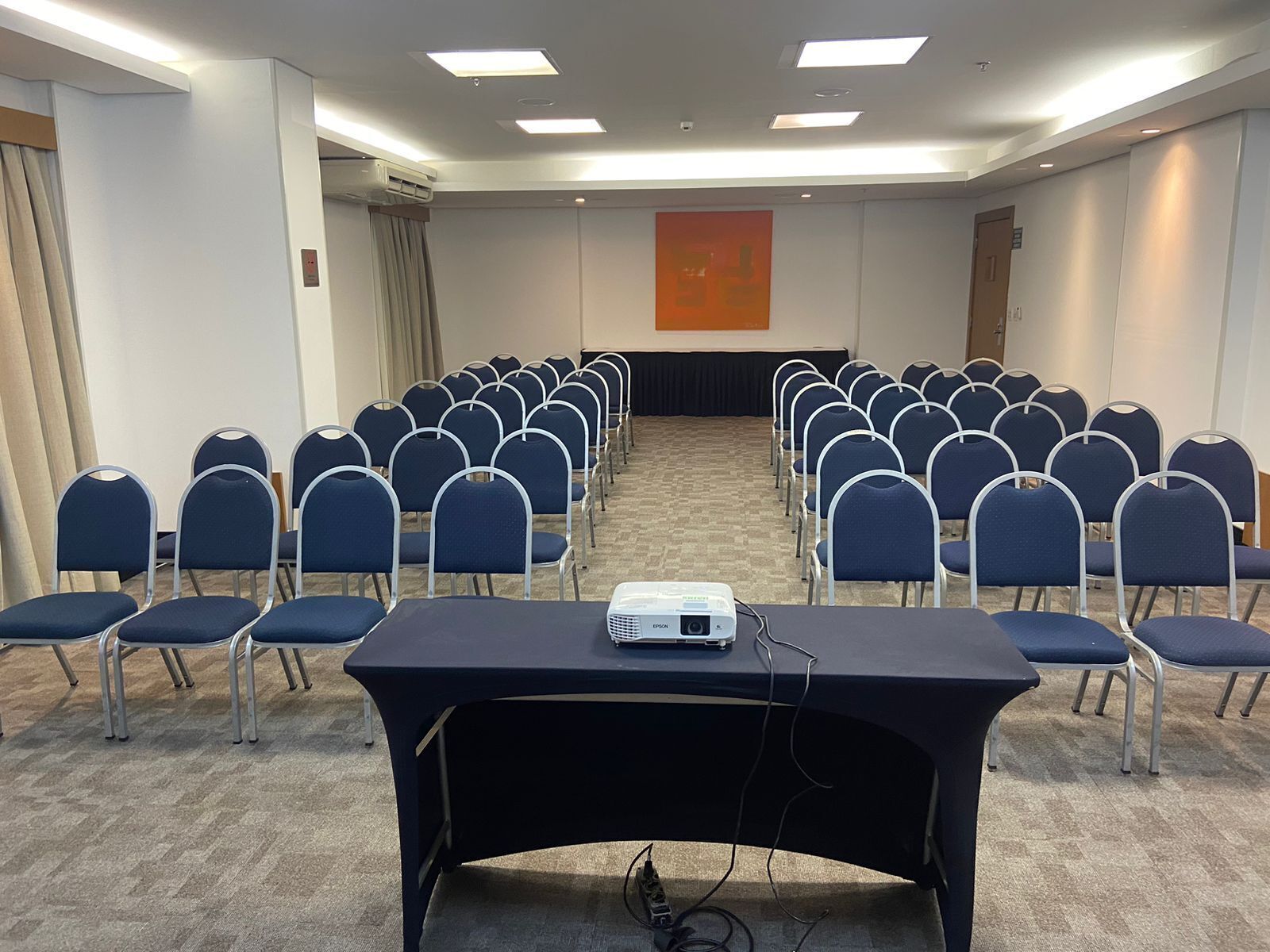 Uma sala de conferência com fileiras de cadeiras, um projetor sobre uma mesa e piso carpeteado.