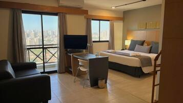 Quarto de hotel com cama, escrivaninha, cadeira, televisão e amplas janelas com vista para a cidade.