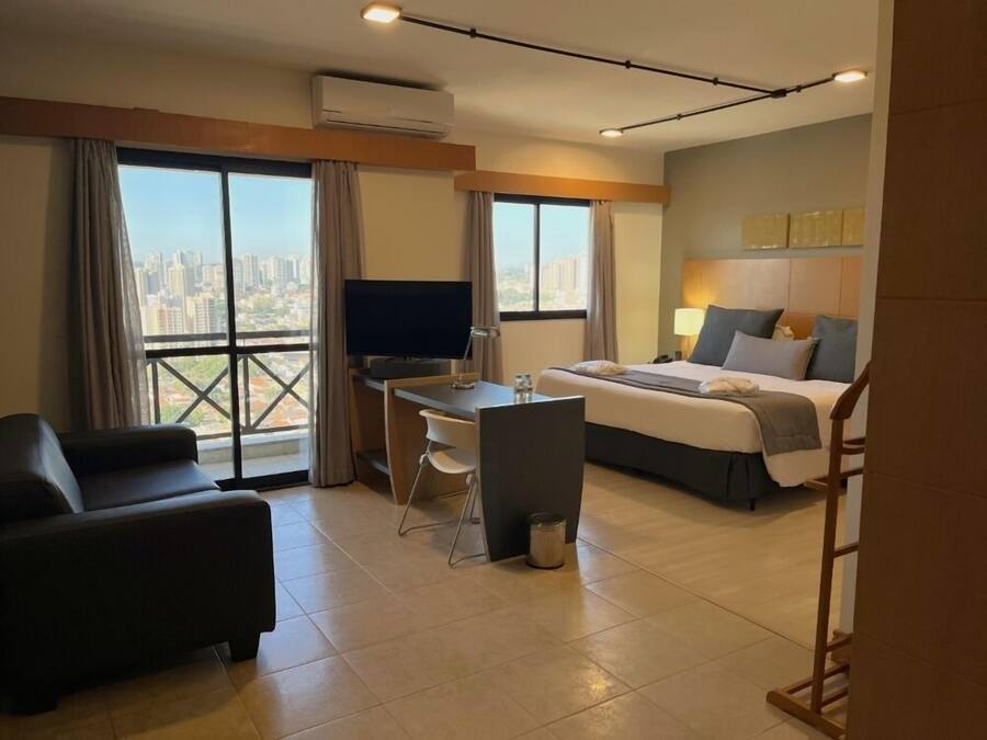 Quarto de hotel com cama, escrivaninha, cadeira, televisão e amplas janelas com vista para a cidade.