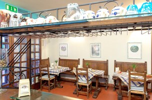 Uma área de refeições com mesas e bancos de madeira, uma escada e uma exposição de pratos de cerâmica na parede.