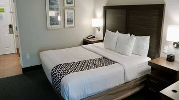 Quarto de hotel com uma cama grande, uma mesa de cabeceira com abajur e um telefone fixado na parede. Há quadros enquadrados na parede e uma porta à esquerda.