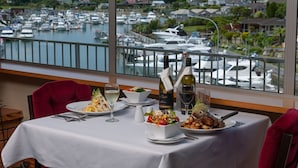 Mesa de jantar com vista para uma marina e barcos, disposta com pratos de comida, copos de vinho e talheres.