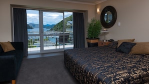 Quarto de hotel com uma cama grande, uma cadeira, uma planta em vaso, uma mesa de cabeceira e vista para uma marina e montanhas.