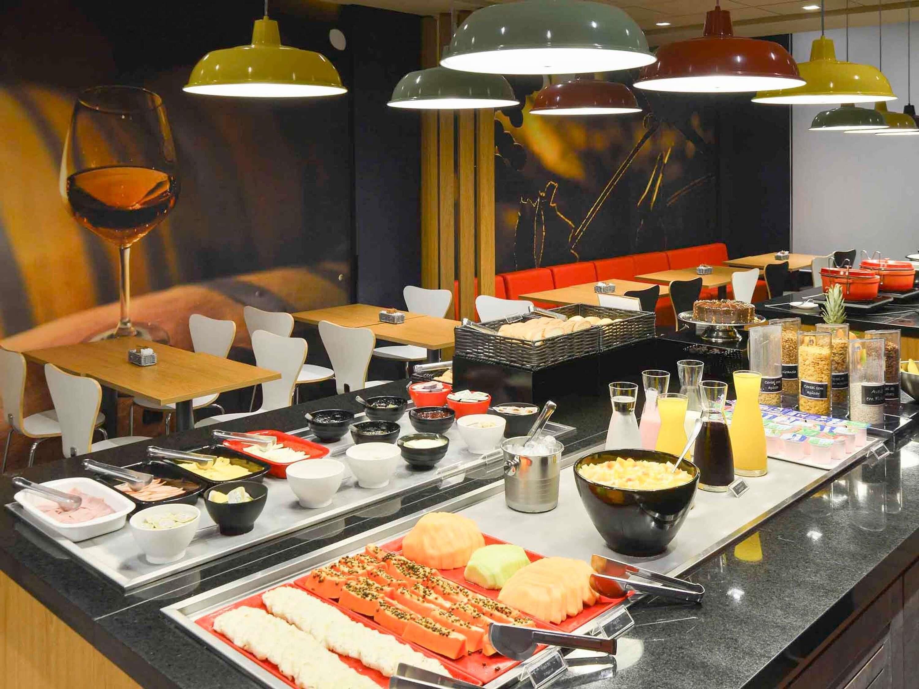 Um buffet com diversos alimentos, acompanhamentos, uma taça de vinho, várias mesas e cadeiras.