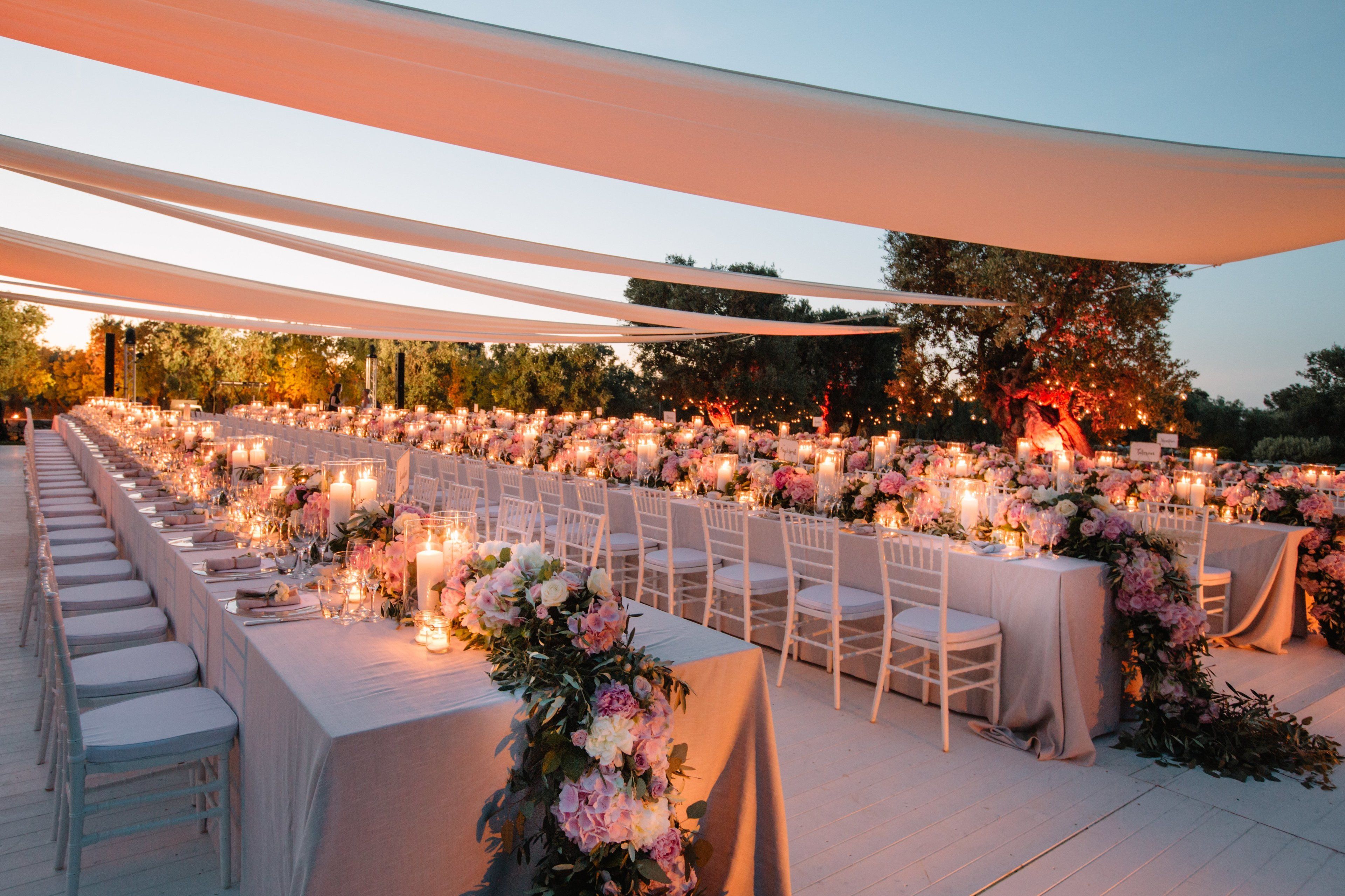 Um salão de banquetes ao ar livre, com mesas compridas, cadeiras brancas e arranjos florais.