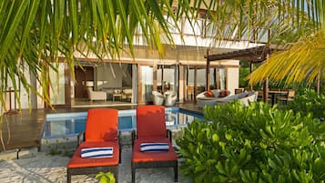 Uma área na beira da piscina com duas espreguiçadeiras vermelhas, uma piscina com proteção de vidro e um edifício moderno com grandes janelas.