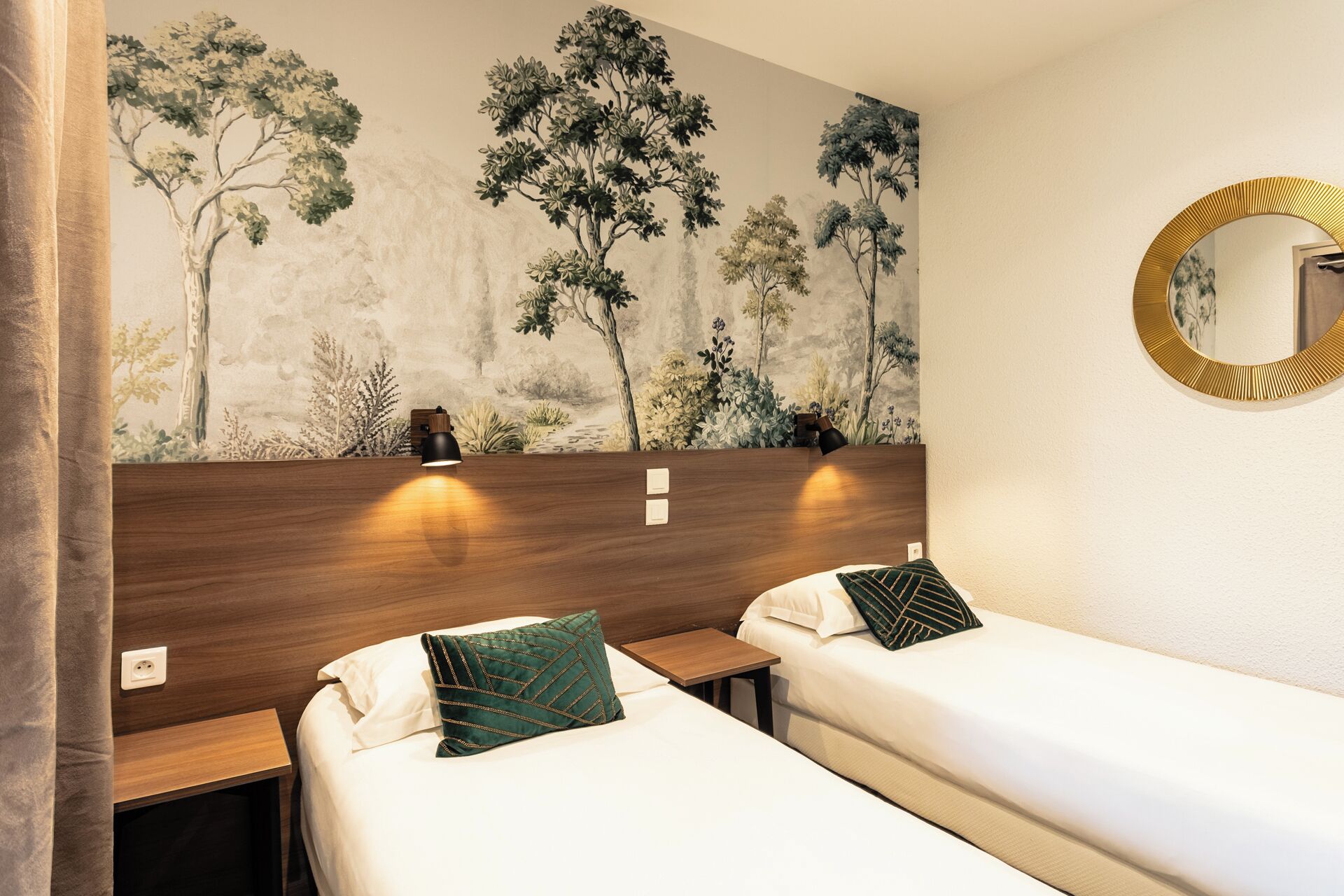 Quarto de hotel com duas camas de solteiro, uma mesa de cabeceira, uma luminária na parede e uma grande pintura de árvores.
