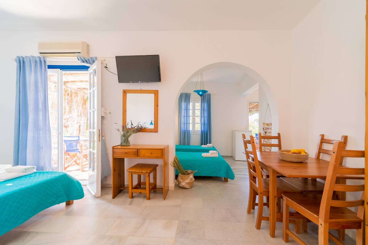 Quarto com duas camas, uma escrivaninha, uma mesa de jantar com cadeiras, uma televisão e uma porta em arco.