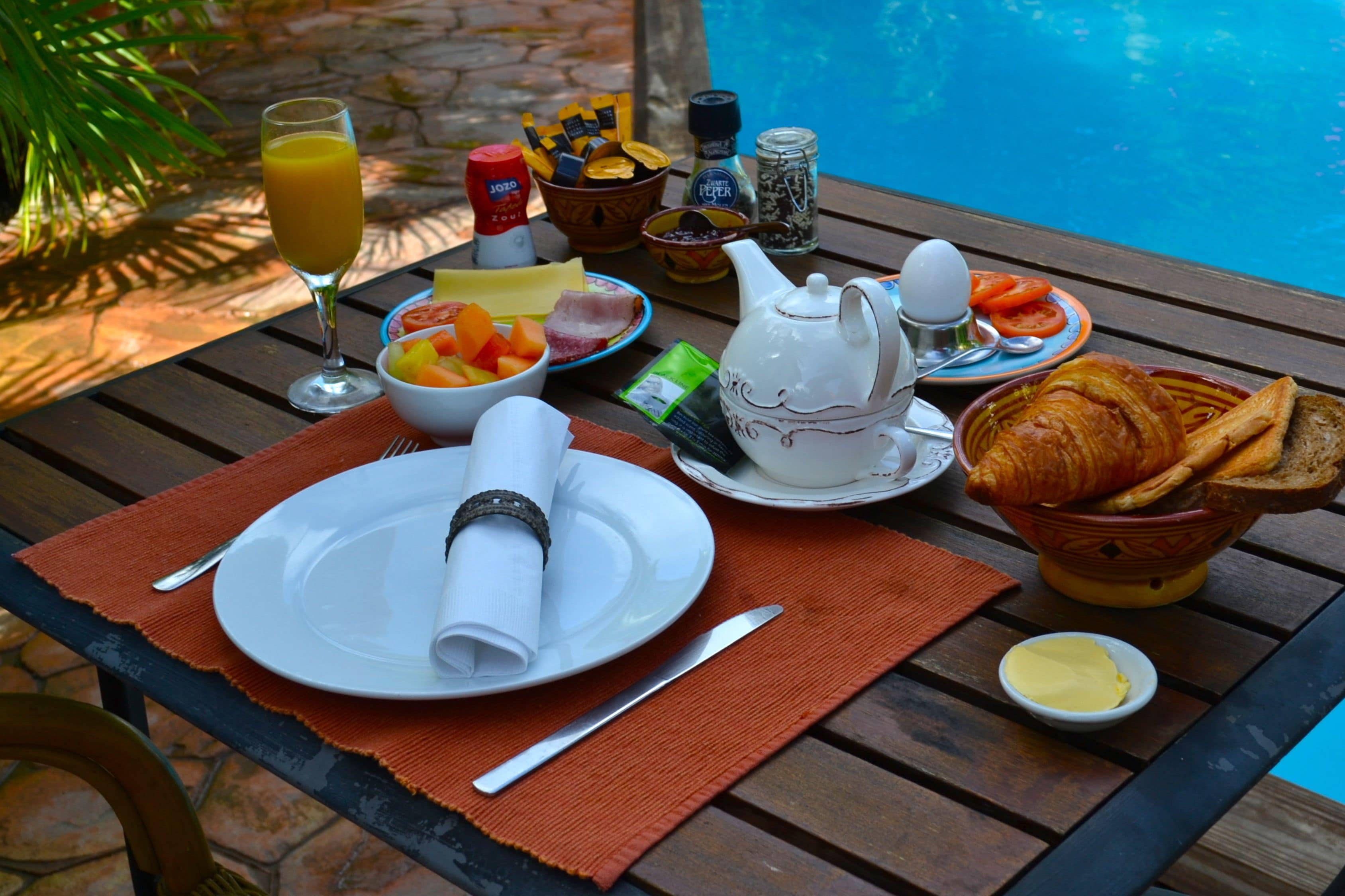 Disposição para o pequeno-almoço com um croissant, torradas, fruta e um bule de chá sobre uma mesa de madeira junto a uma piscina.