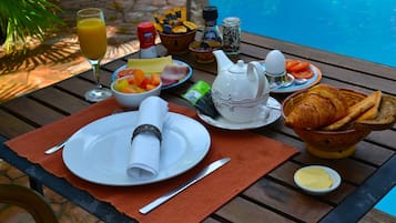 Disposição para o pequeno-almoço com um croissant, torradas, fruta e um bule de chá sobre uma mesa de madeira junto a uma piscina.