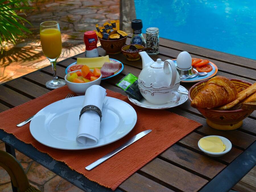 Disposição para o pequeno-almoço com um croissant, torradas, fruta e um bule de chá sobre uma mesa de madeira junto a uma piscina.