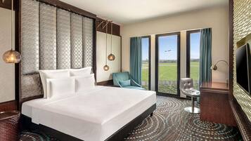 Quarto de hotel com uma cama grande, uma cadeira, uma televisão e vista para o aeroporto.