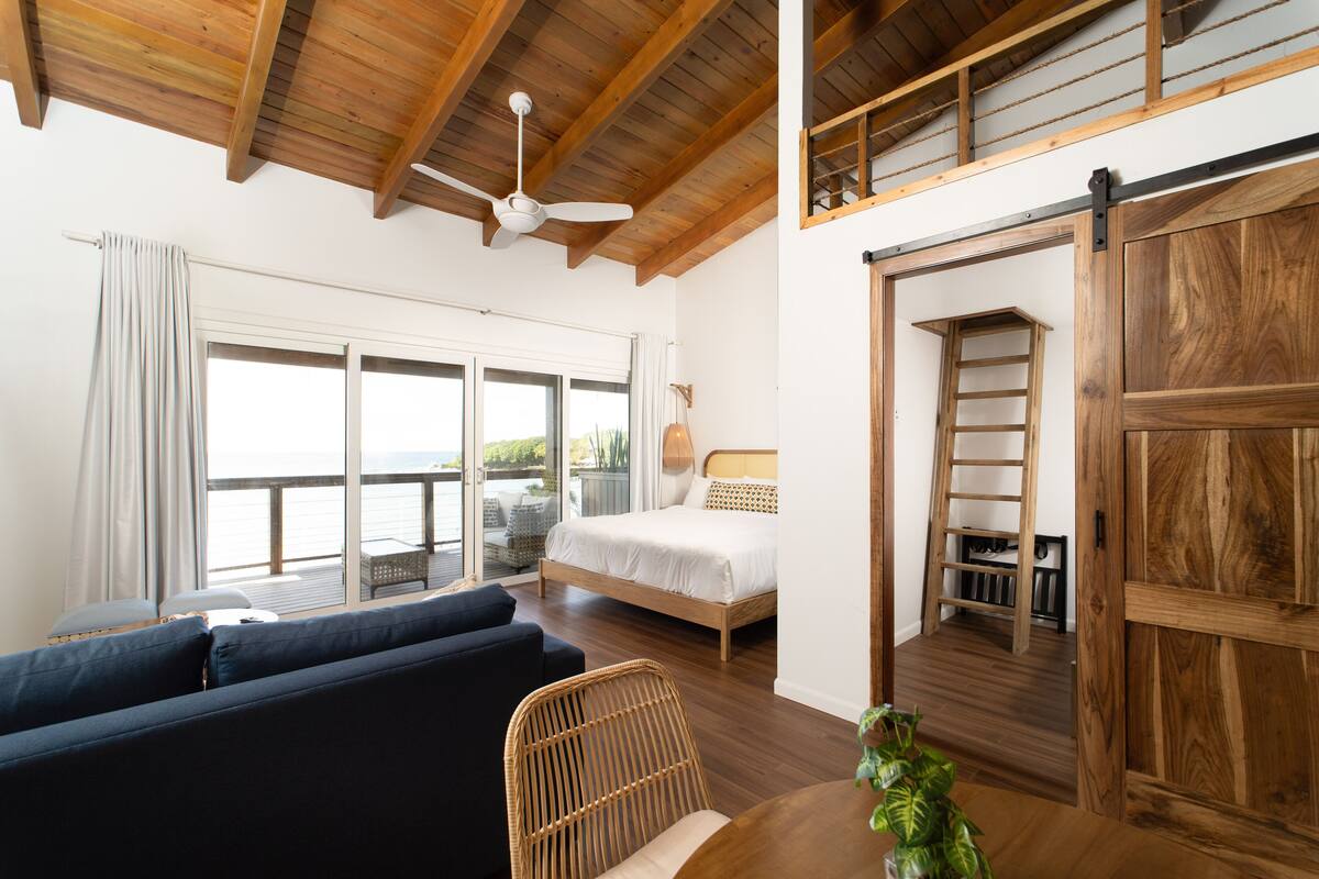 Quarto com cama, ventilador de teto, piso de madeira, varanda com vista e porta de correr.