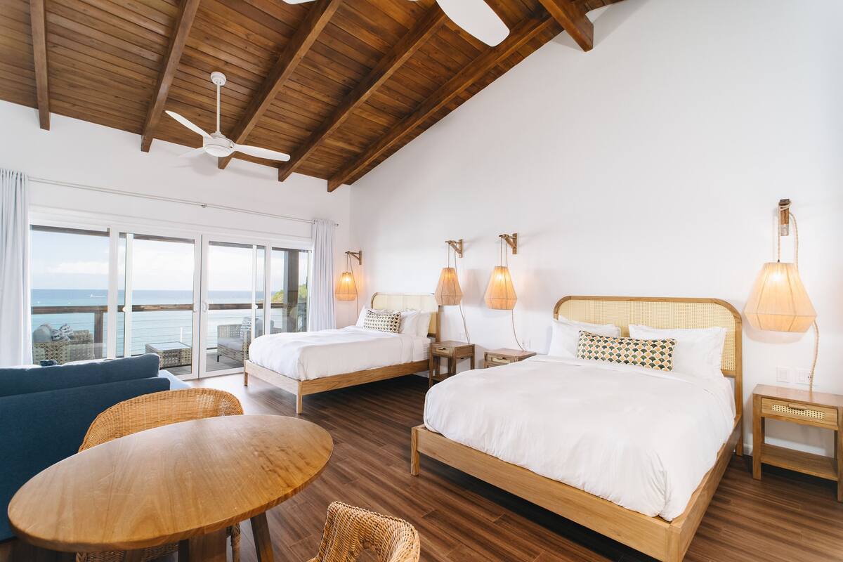 Quarto de hotel com duas camas, ventilador de teto, janelas amplas com vista e varanda.