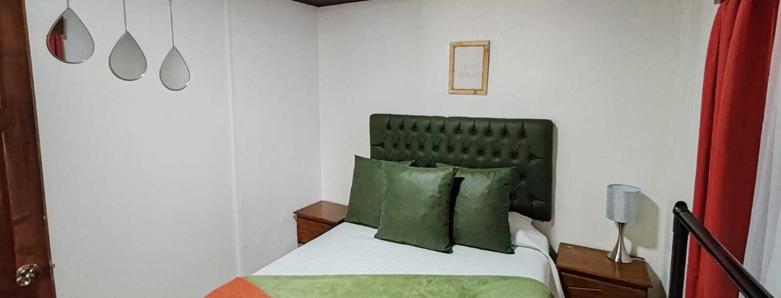 Quarto com cabeceira estofada verde, colcha branca e duas almofadas verdes. Tem uma pequena mesa de cabeceira de madeira com uma luminária ao lado da cama.