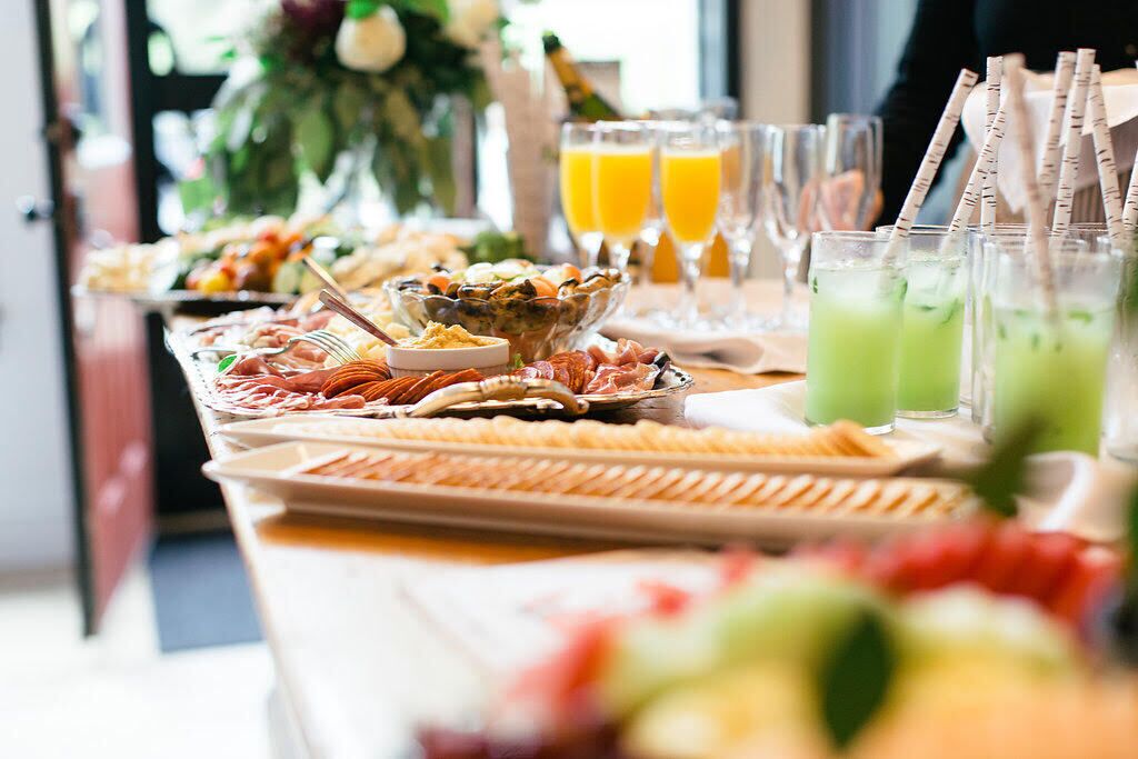 Uma mesa de buffet com diversos alimentos e bebidas, incluindo uma travessa de frutos do mar, um prato de bolachas e copos de suco de laranja e bebidas verdes.