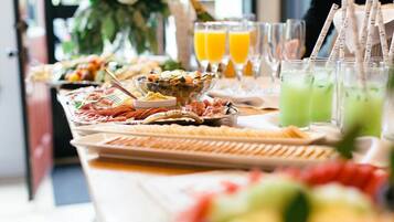 Uma mesa de buffet com diversos alimentos e bebidas, incluindo uma travessa de frutos do mar, um prato de bolachas e copos de suco de laranja e bebidas verdes.