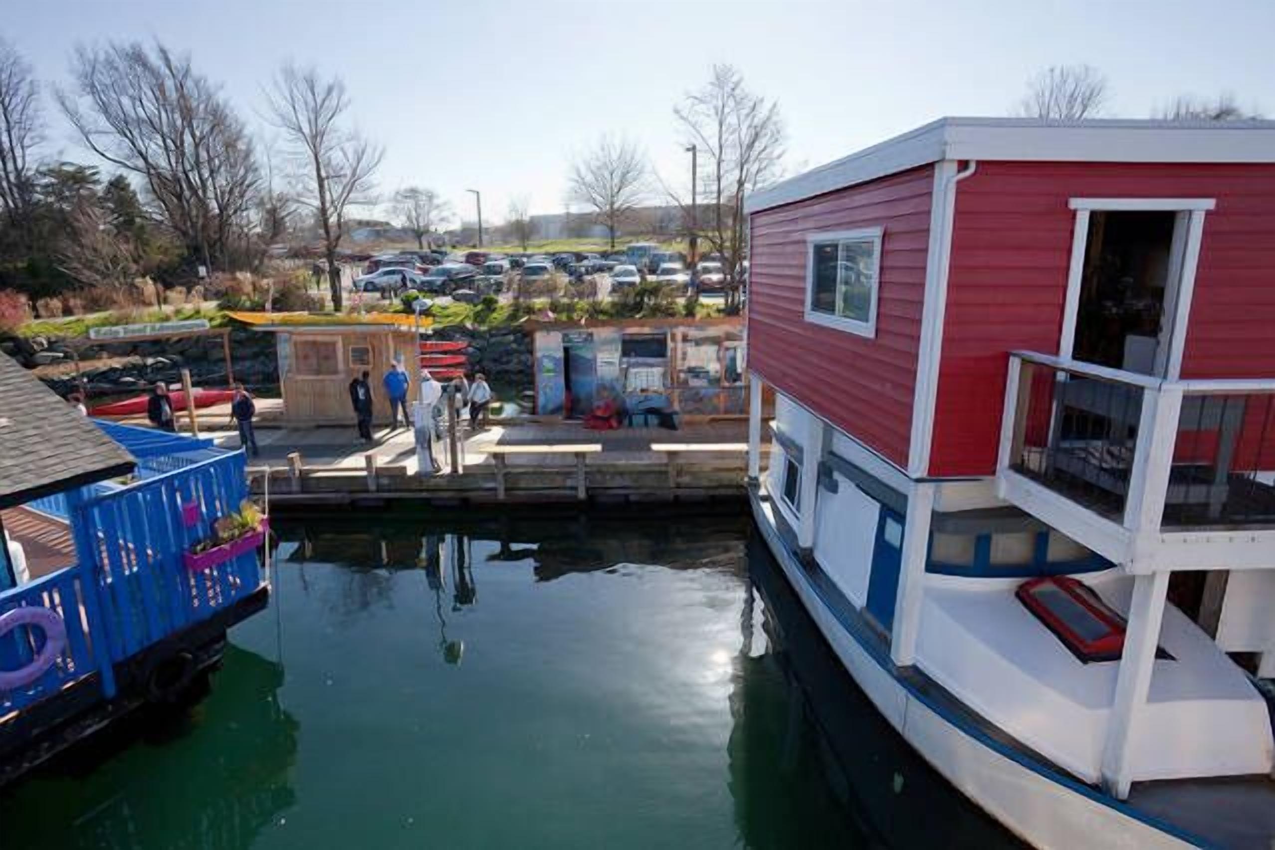 Uma casa-barco atracada, com um pequeno deque e área coberta, rodeada por águas calmas e carros estacionados ao fundo.