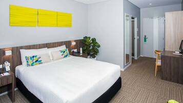 Quarto de hotel com uma cama grande, mesas de cabeceira, uma escrivaninha, um espelho e um vaso de plantas.