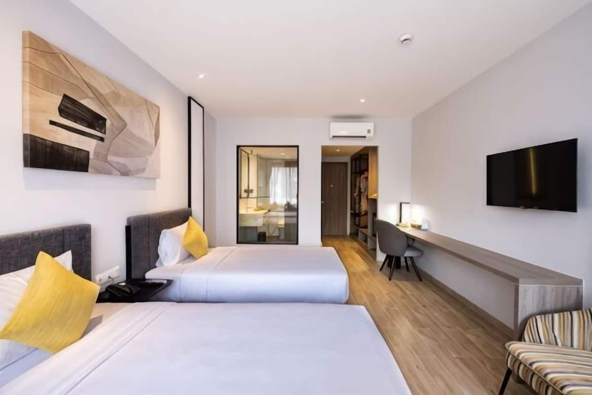 Quarto de hotel com duas camas, uma escrivaninha, uma cadeira, televisão e banheiro visível por uma porta de vidro.