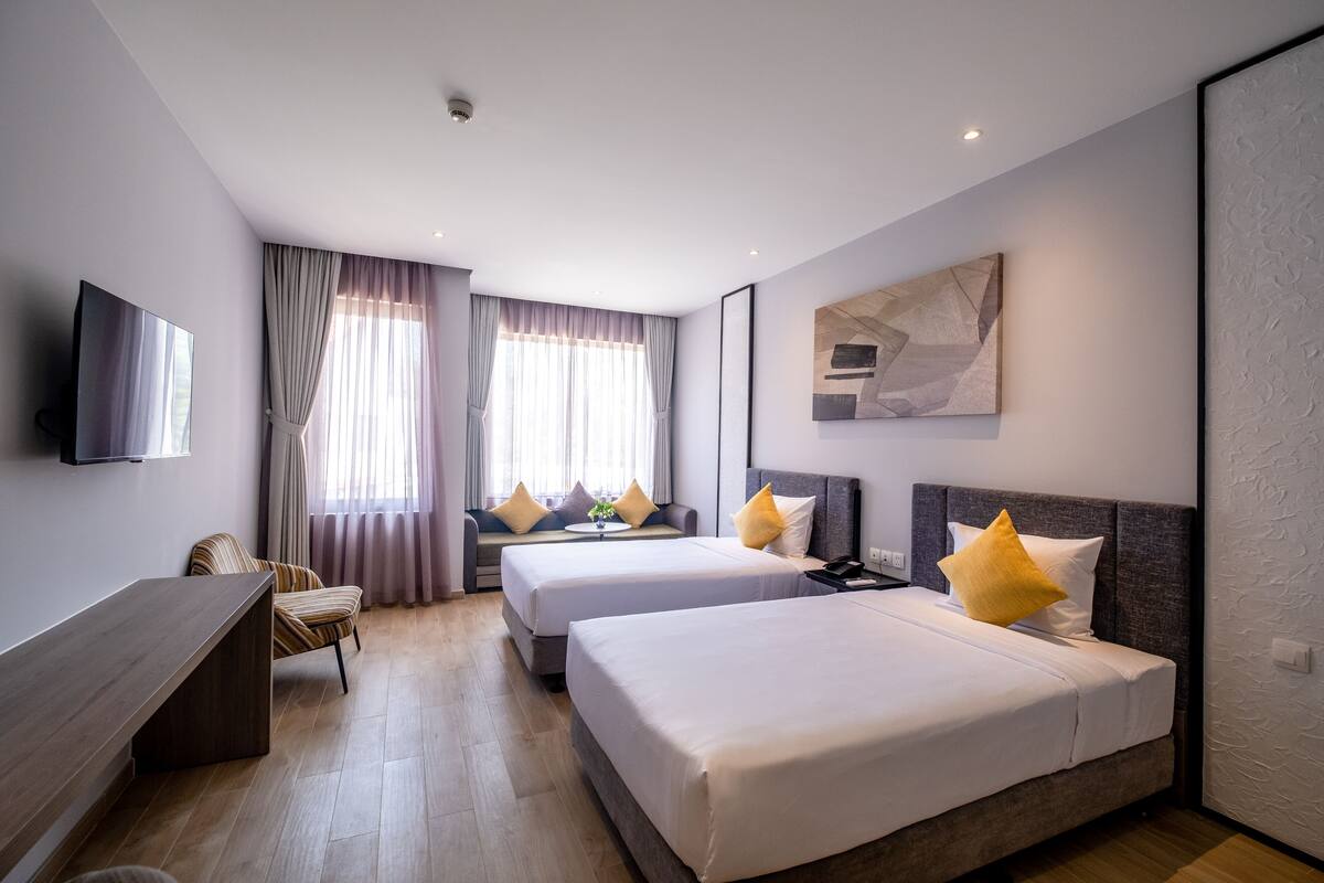 Quarto de hotel com duas camas, uma escrivaninha, televisão e janela com cortinas.