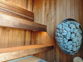 Uma sauna de madeira com um banco, uma cesta de metal cheia de pedras e piso de madeira.