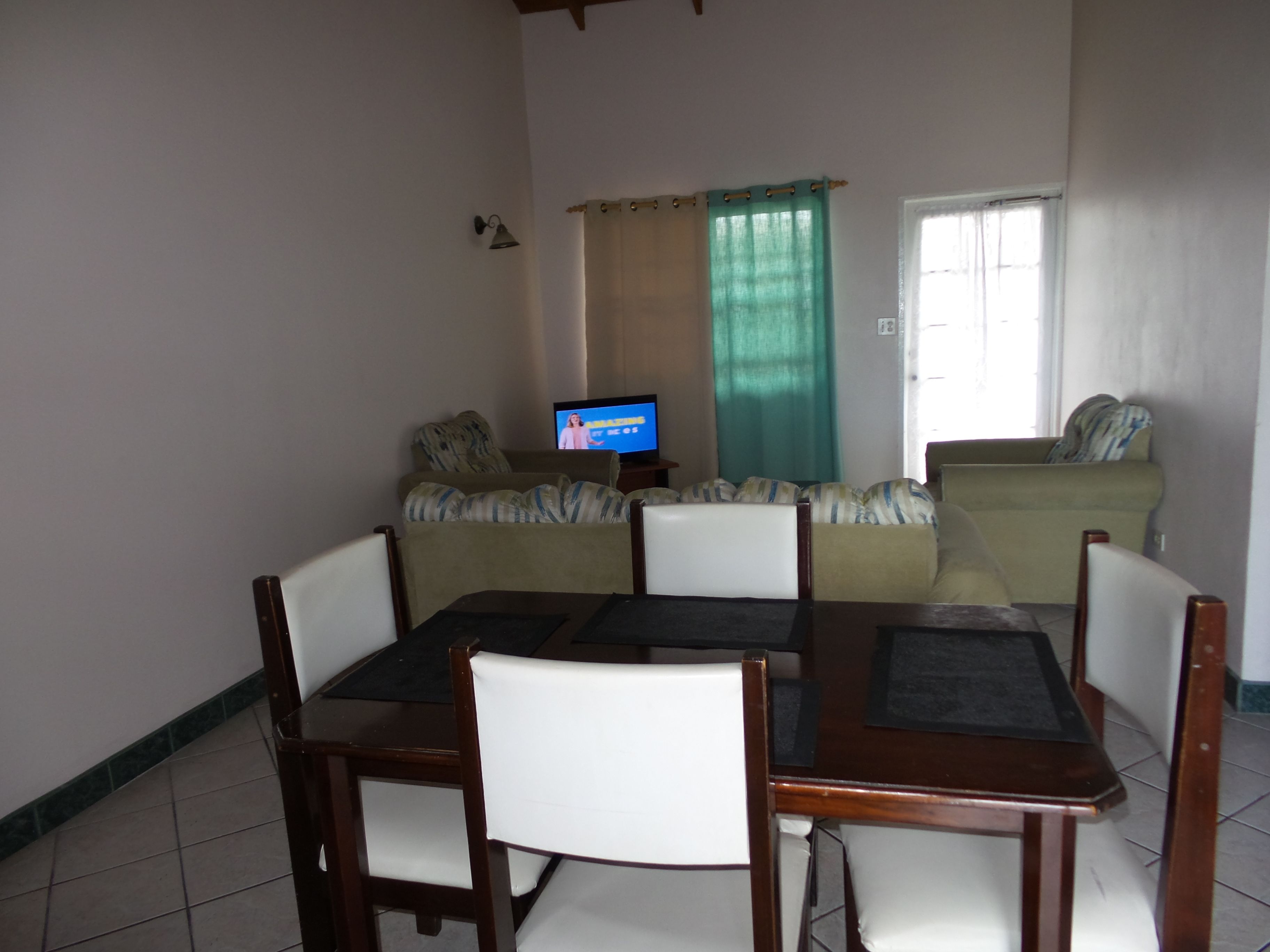 Uma área de refeições com uma mesa e cadeiras, uma televisão ao fundo e uma janela com cortinas.
