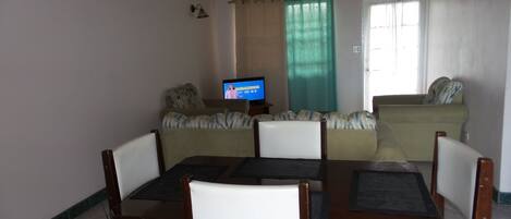 Uma área de refeições com uma mesa e cadeiras, uma televisão ao fundo e uma janela com cortinas.