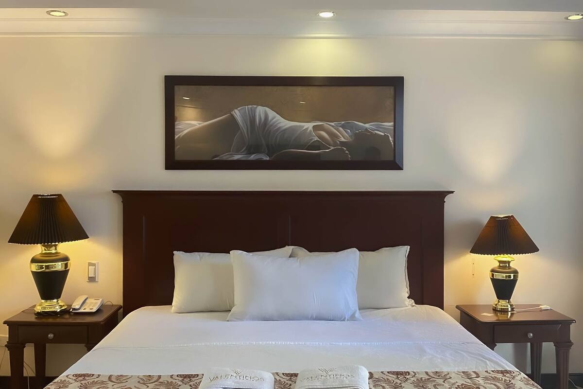 Quarto de hotel com uma cama grande, duas mesas de cabeceira com abajures, uma obra de arte emoldurada acima da cama e uma cama bem arrumada com toalhas.