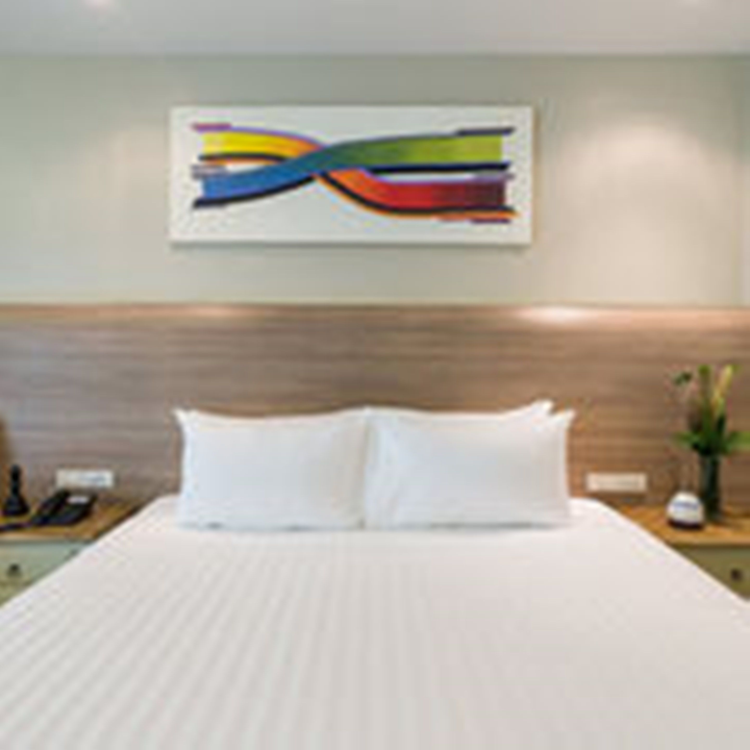 Quarto de hotel com cama bem arrumada, mesa de cabeceira com luminária e pintura abstrata na parede.