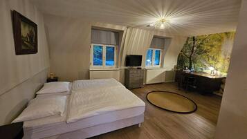 Quarto com cama, escrivaninha, cadeira, televisão e janela com vista para árvores.
