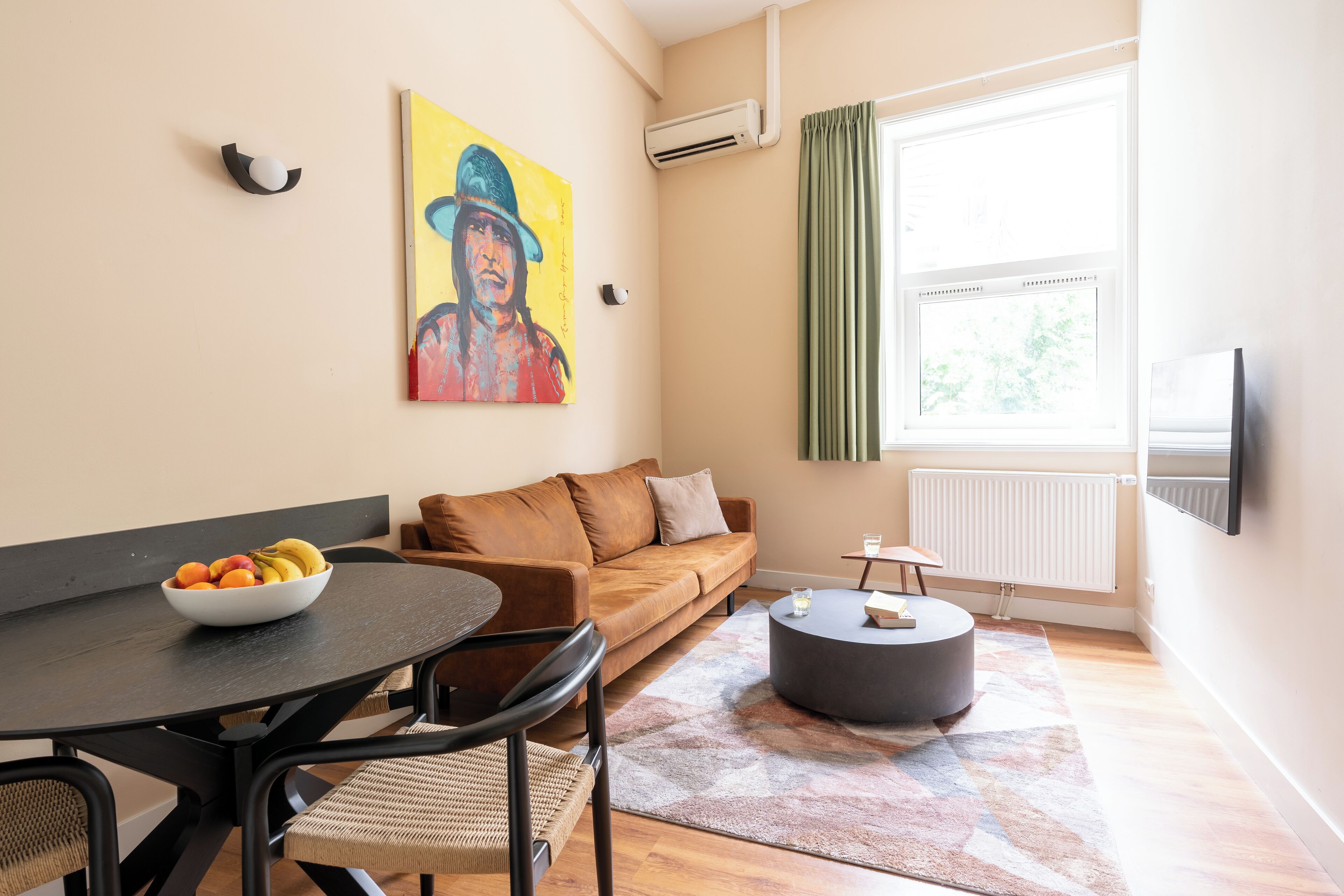 Uma sala de estar com um sofá marrom, uma mesa de centro redonda, uma mesa de jantar com cadeiras, um quadro na parede e uma janela com cortinas.