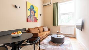 Uma sala de estar com um sofá marrom, uma mesa de centro redonda, uma mesa de jantar com cadeiras, um quadro na parede e uma janela com cortinas.