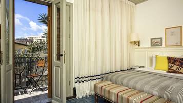 Quarto de hotel com cama, um banquinho, uma cadeira, janela com cortinas, abajur e um quadro na parede.