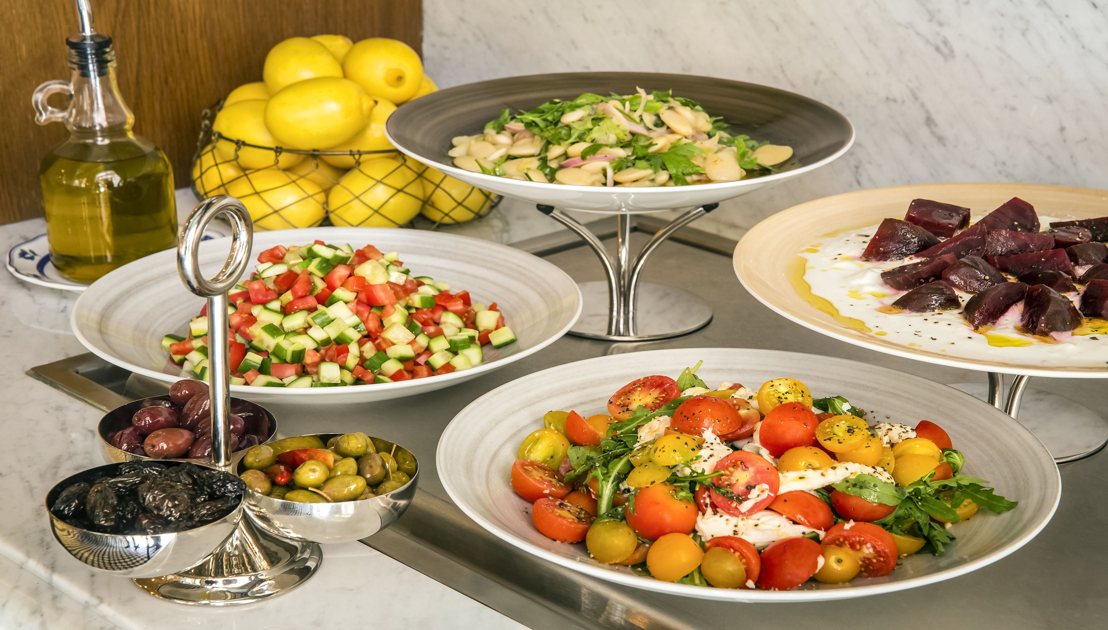 Diversos pratos em uma mesa, incluindo saladas, um recipiente com azeitonas e uma garrafa de azeite.