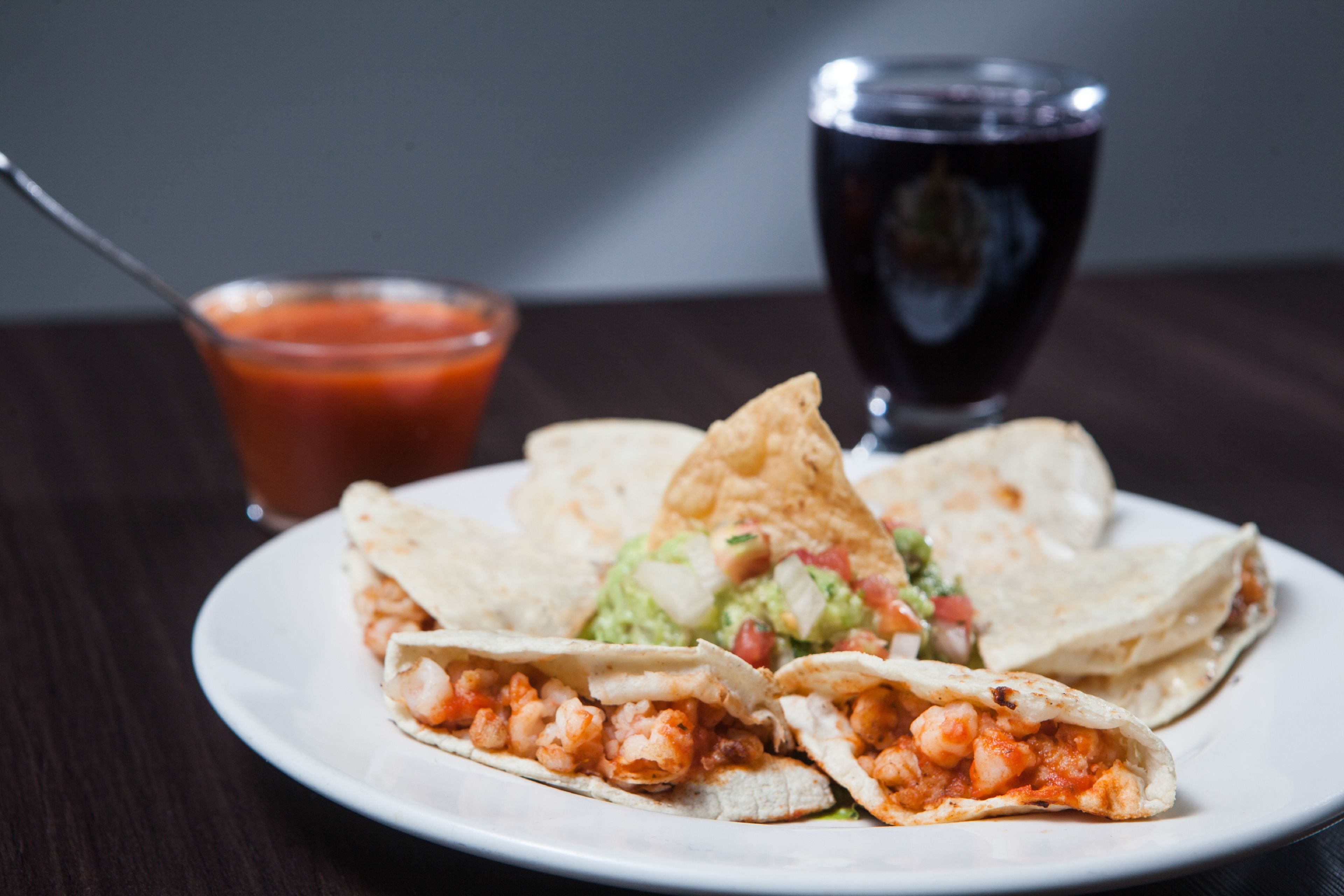 Um prato com três tacos, um copo de bebida escura e uma pequena tigela de molho vermelho.