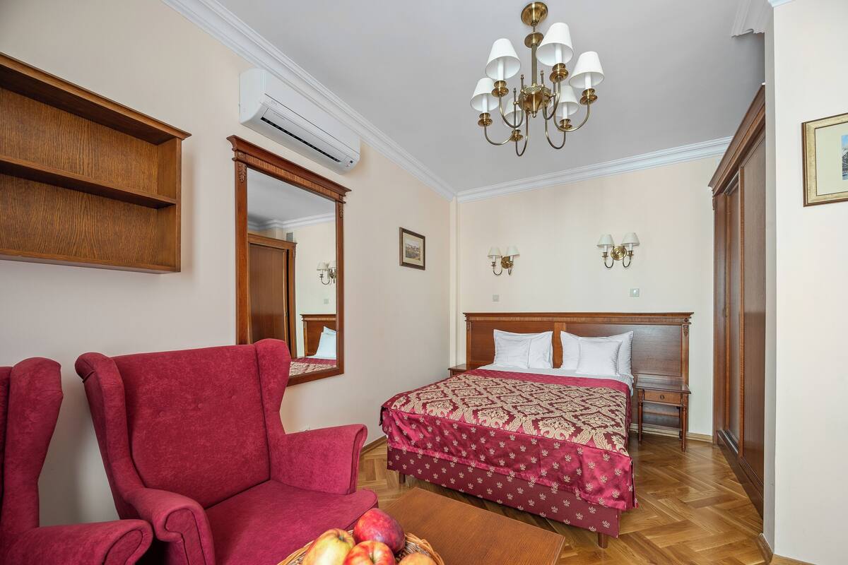 Quarto de hotel com cama de madeira, uma poltrona vermelha, piso de madeira, um candelabro e um espelho grande.