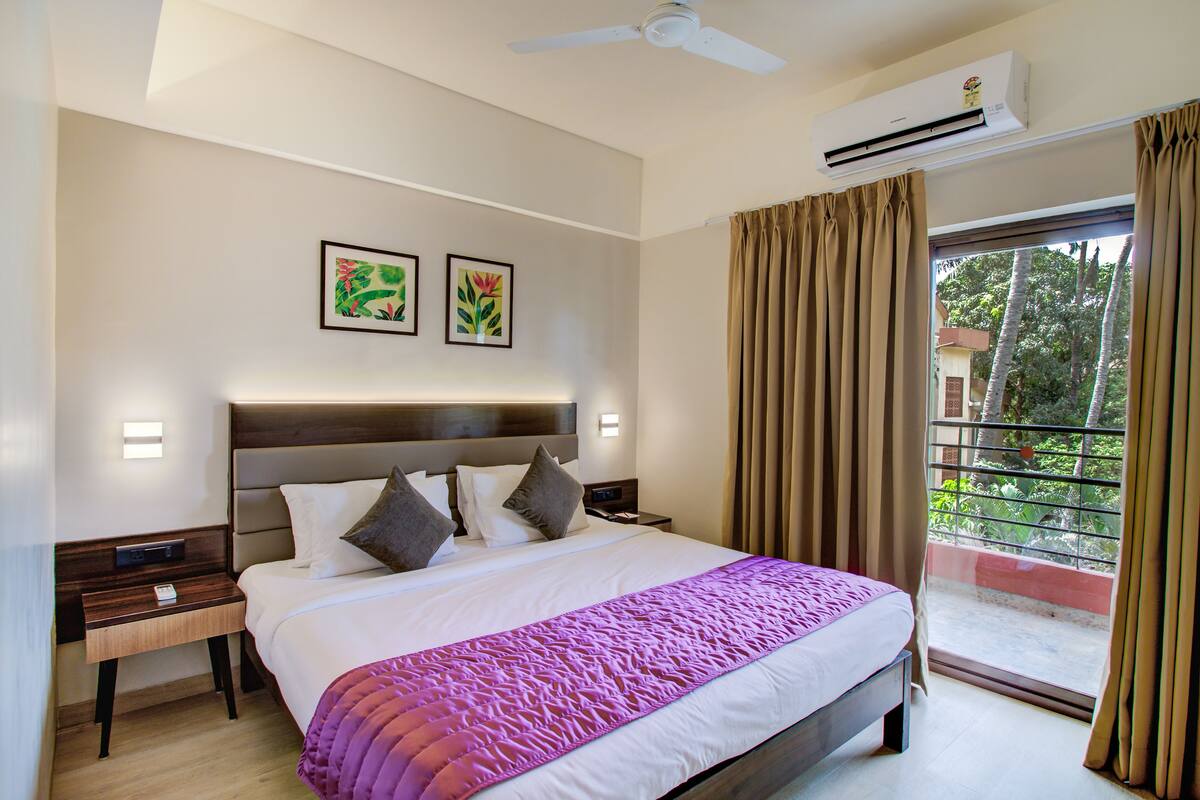 Quarto de hotel com cama, mesas de cabeceira, ventoinha no teto, ar condicionado e janela com cortinas.