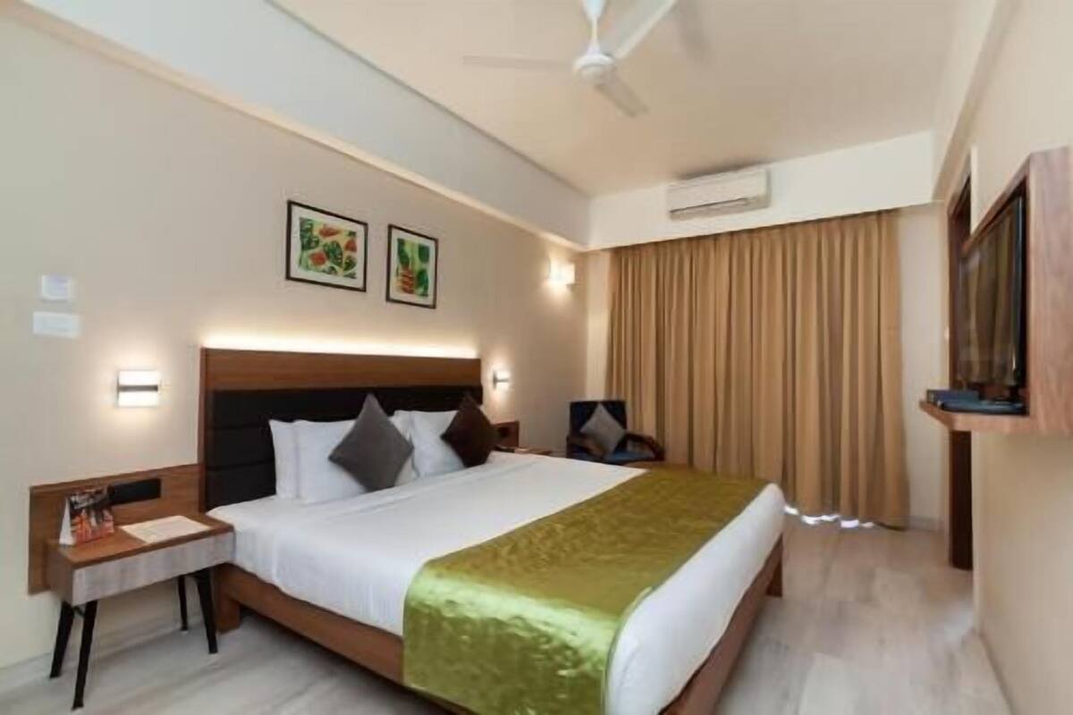 Quarto de hotel com cama, mesas de cabeceira, escrivaninha, ventoinha no teto e janela com cortinas.