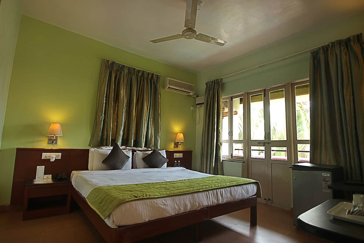 Quarto de hotel com uma cama, mesas de cabeceira, escrivaninha, ventoinha no teto, ar condicionado e janela com cortinas.