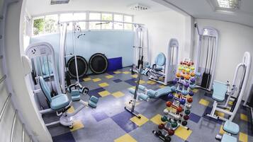 Uma academia com equipamentos de exercícios, piso quadriculado azul e amarelo, e janelas amplas.