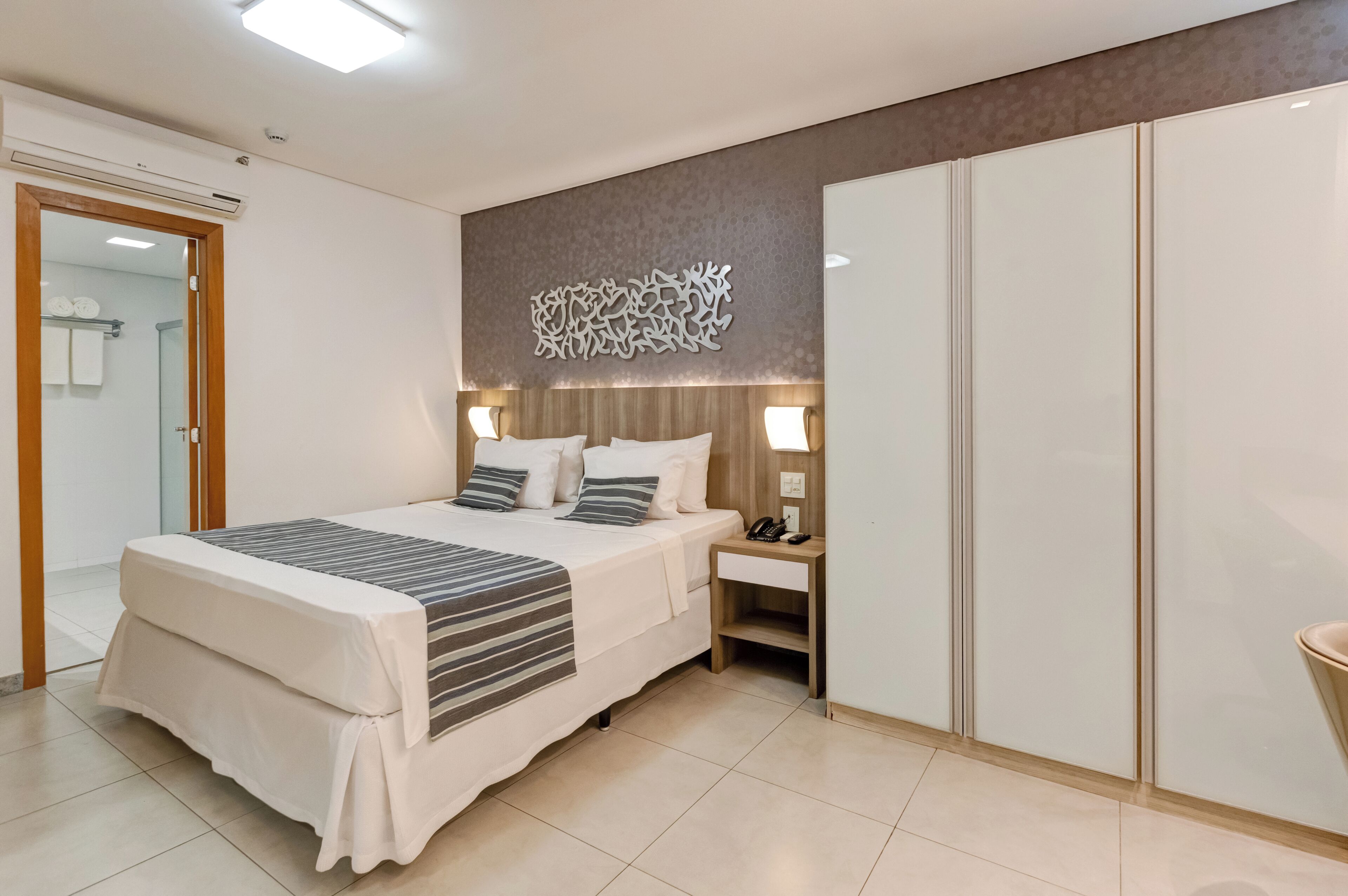 Quarto moderno com cama grande, luminárias de cabeceira, guarda-roupa e um detalhe decorativo na parede.