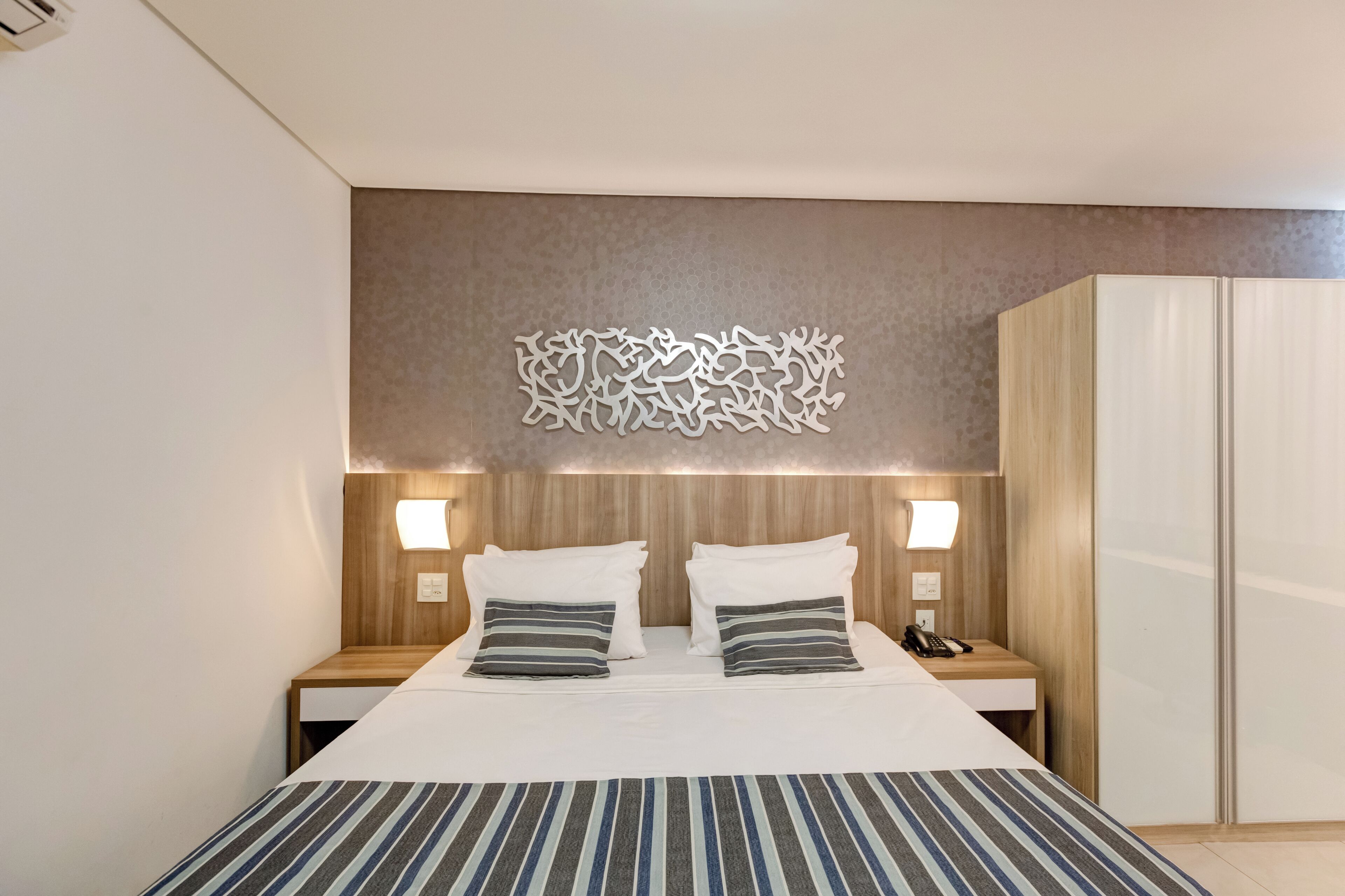 Quarto de hotel moderno com uma cama grande, duas mesas de cabeceira, um painel decorativo na parede e um guarda-roupa.