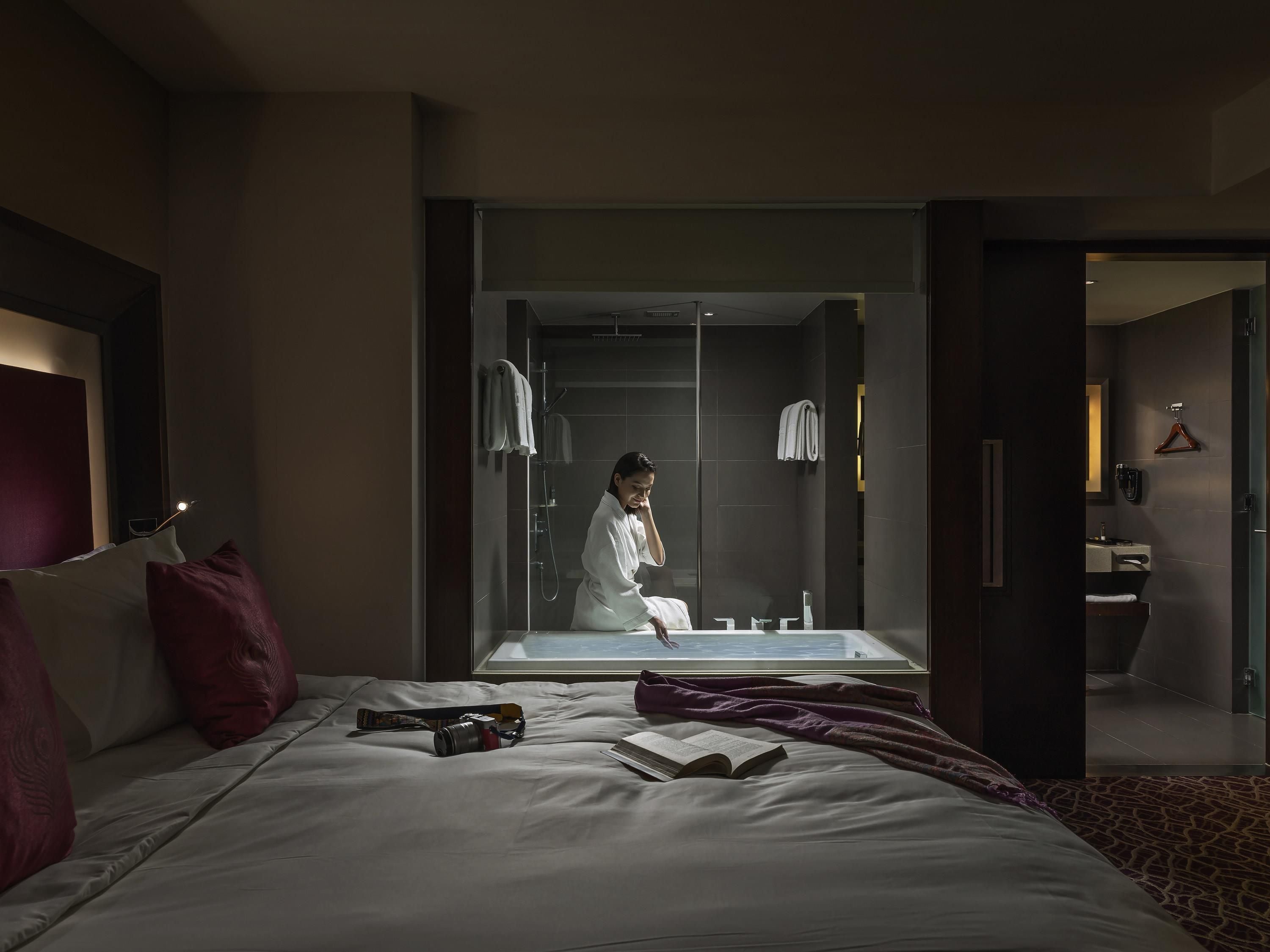 Quarto de hotel com uma cama, banheiro visível através de um painel de vidro e um porta-toalhas.