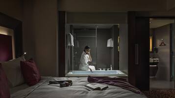 Quarto de hotel com uma cama, banheiro visível através de um painel de vidro e um porta-toalhas.