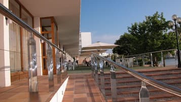 Corredor com piso de azulejos, corrimão de metal e vista para um corpo de água.