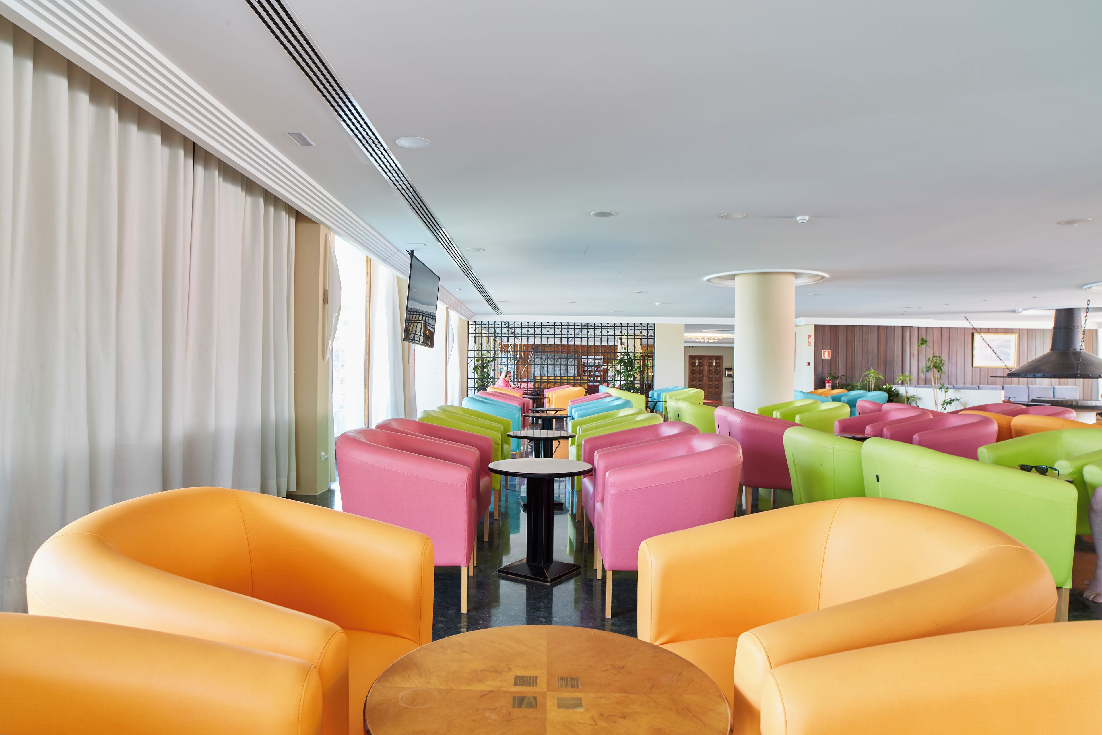 Um saguão de hotel com assentos coloridos, uma mesa central e vista para a área de refeições.